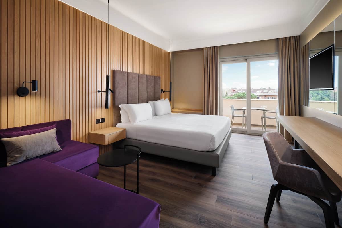 Quarto de hotel moderno com uma cama grande, um sofá roxo, uma pequena mesa e uma escrivaninha. O quarto possui painéis de madeira nas paredes e uma grande janela com vista.