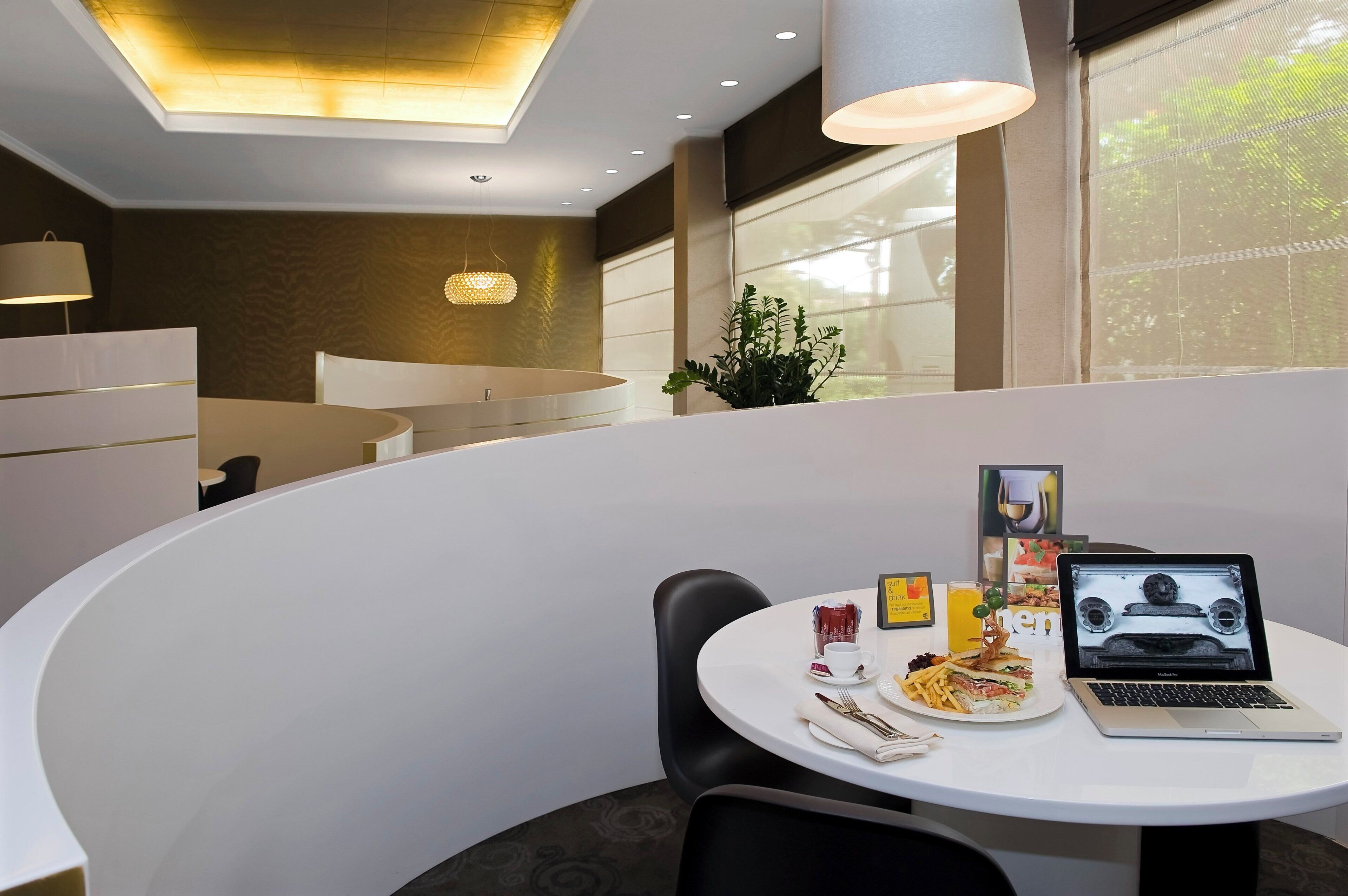 Mesa de jantar moderna e curva, com um laptop, um copo de suco e um sanduíche.