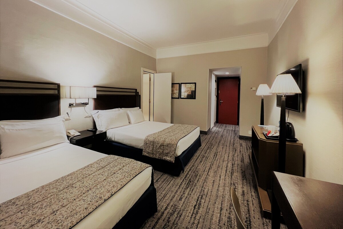Quarto de hotel com duas camas, uma escrivaninha, uma cadeira, uma TV e uma porta que dá para outro espaço.