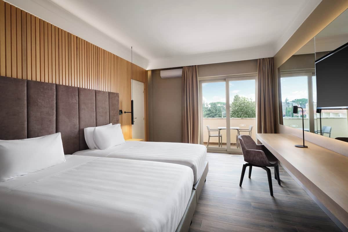 Quarto de hotel com duas camas, uma mesa, uma cadeira, uma janela grande com vista para vegetação e uma televisão de tela plana.