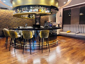 Uma área de bar com balcão circular, vários bancos e um espaço de estar com sofá e almofadas estampadas.