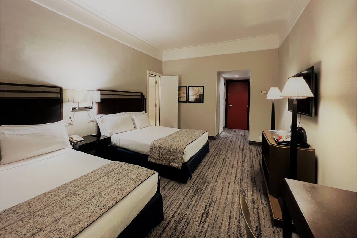 Quarto de hotel com duas camas, uma escrivaninha, uma cadeira, uma TV e uma porta que dá para outro espaço.