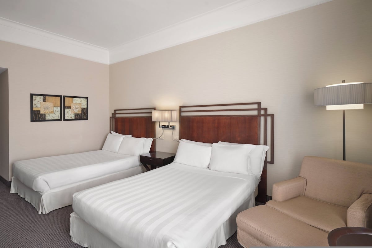 Quarto de hotel com duas camas, um sofá, uma mesa de cabeceira com abajur e uma obra de arte na parede.