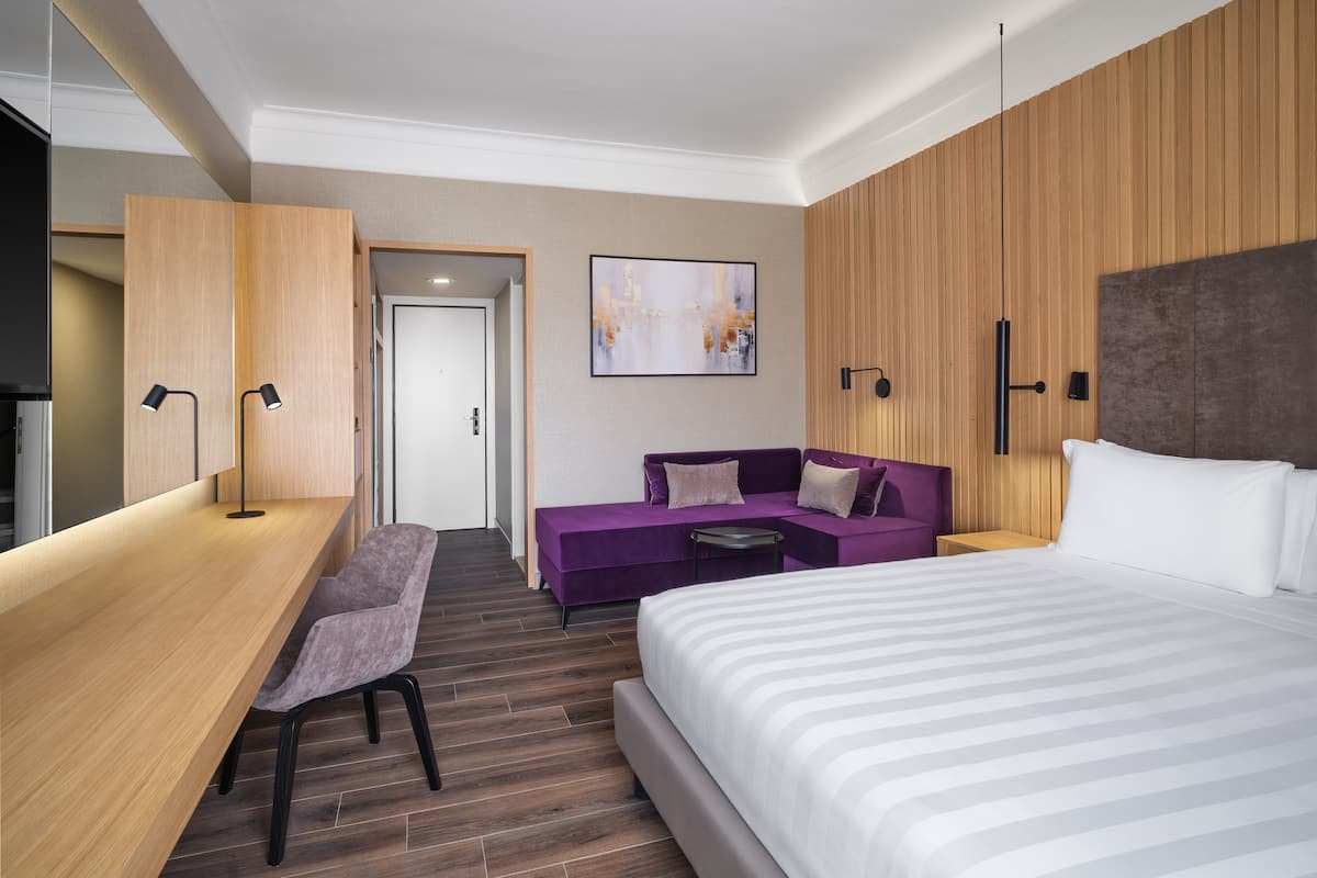 Quarto de hotel moderno com uma cama grande, uma escrivaninha com cadeira, um sofá roxo e uma mesinha.