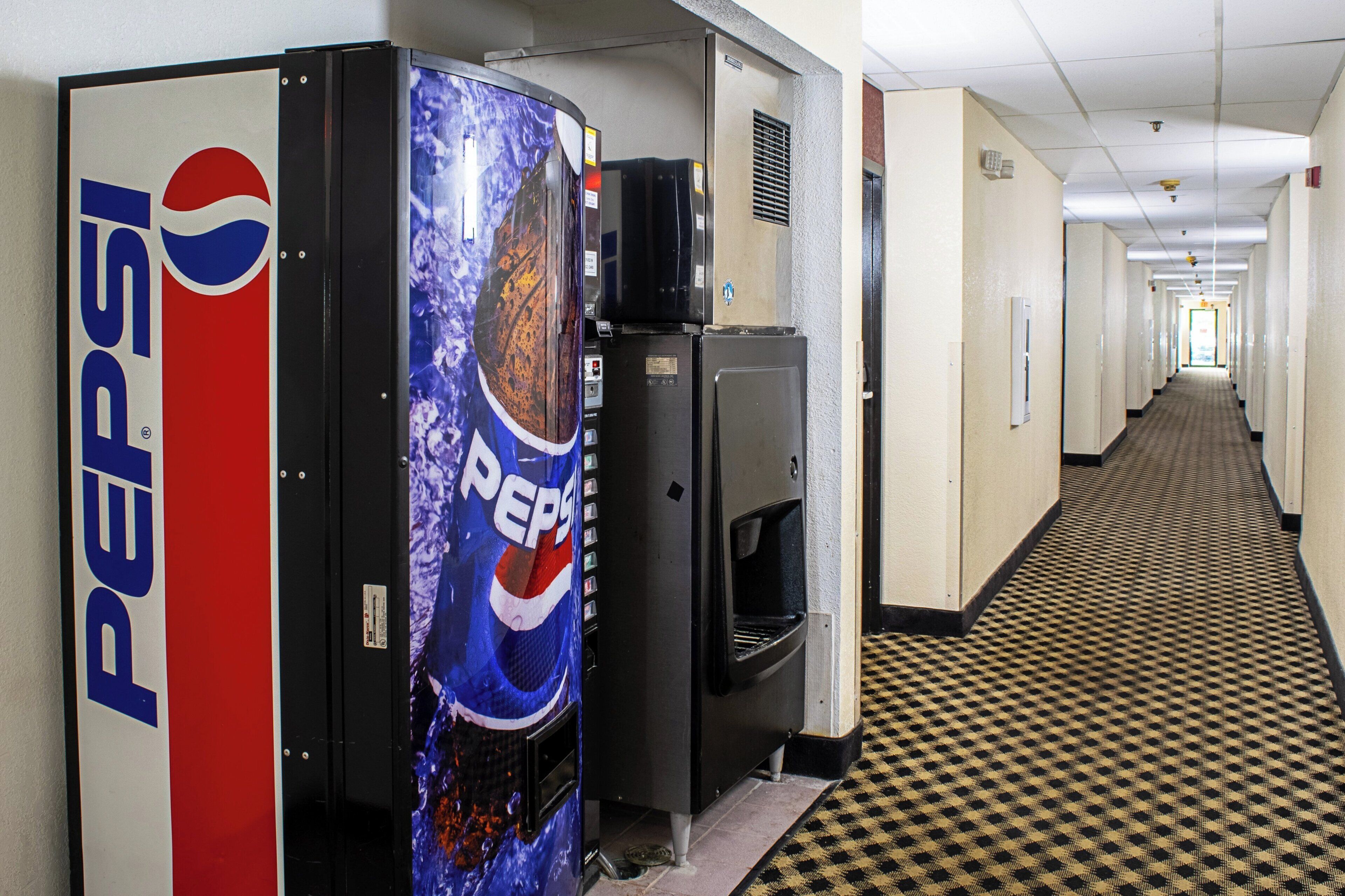 Um corredor com máquinas de venda automática, incluindo uma máquina de Pepsi, e um chão quadriculado.