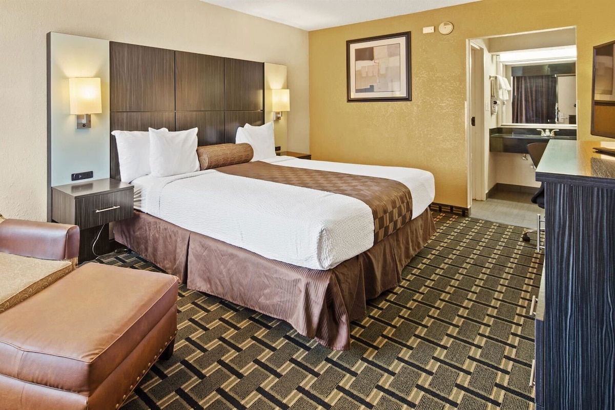 Quarto de hotel com cama, escrivaninha, cadeira, espelho e banheiro visível por uma porta aberta.