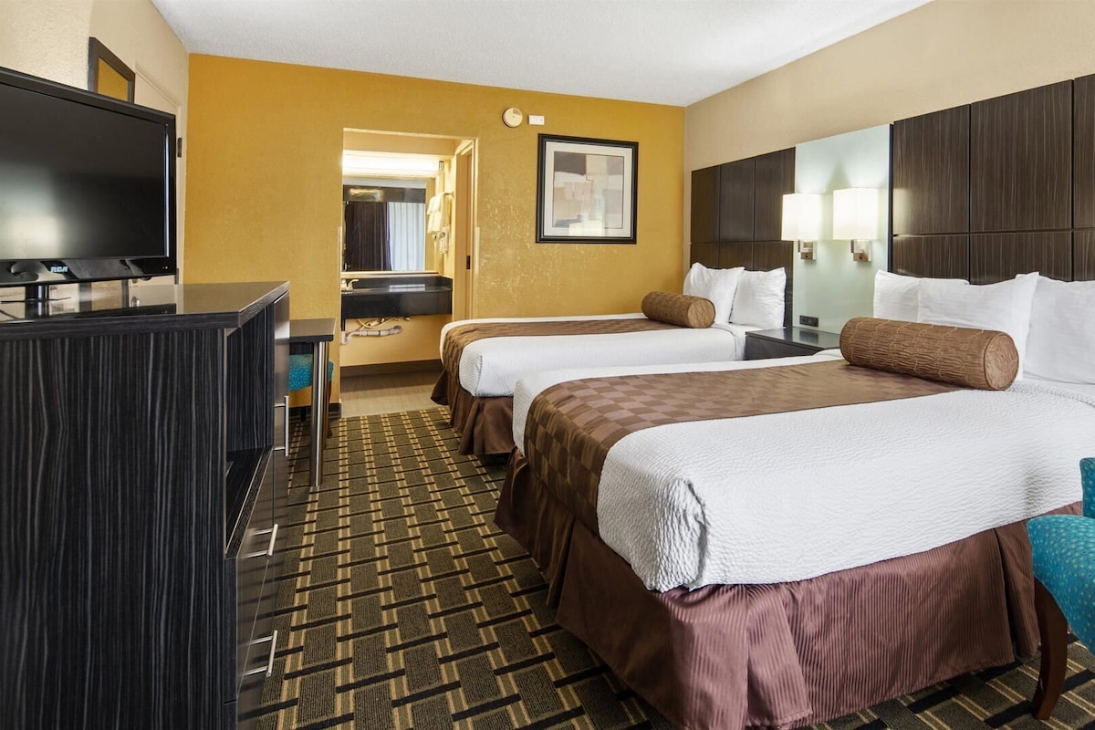 Quarto de hotel com duas camas, uma escrivaninha, uma televisão e um banheiro visível através de uma porta aberta.