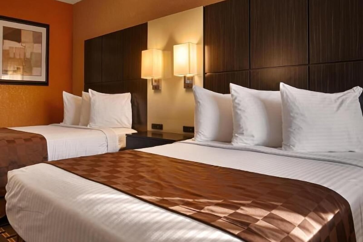 Quarto de hotel com duas camas, uma janela grande e uma luminária fixada na parede.