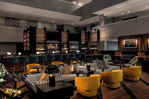 Uma área de bar moderna com sofás xadrez, cadeiras amarelas e um balcão com garrafas.