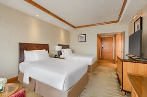 Quarto de hotel com duas camas, uma cabeceira de madeira, uma escrivaninha e uma televisão de tela plana, e uma porta que dá para outro quarto.