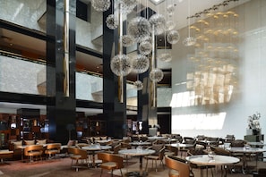 Um amplo hall de hotel com diversas áreas de estar, incluindo mesas redondas e sofás, e um grande candelabro com luminárias esféricas.
