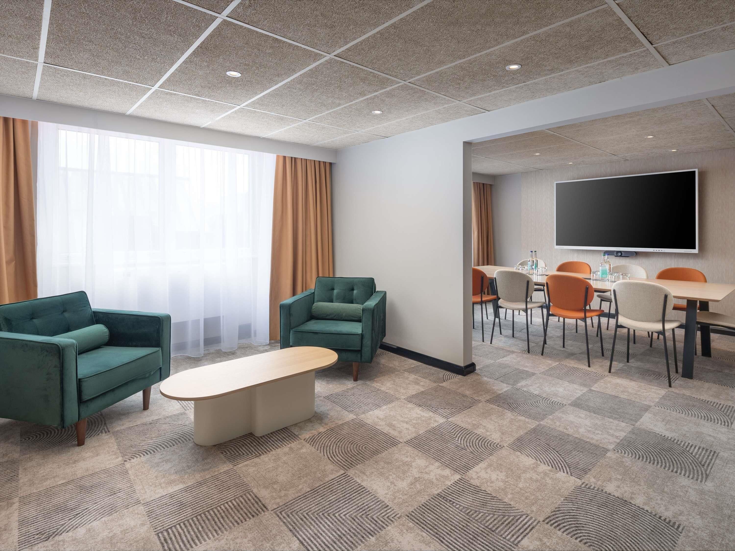 Quarto de hotel com uma televisão grande, um sofá, uma mesa de centro e um espaço de jantar com cadeiras.