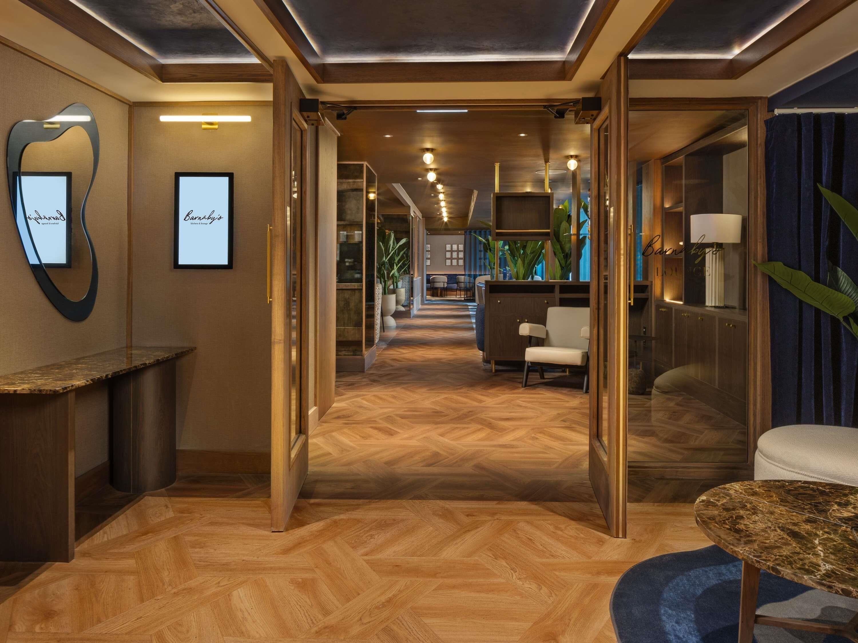 Um hall de hotel com padrão de piso em herringbone, uma recepção e uma área de estar com uma mesa redonda.