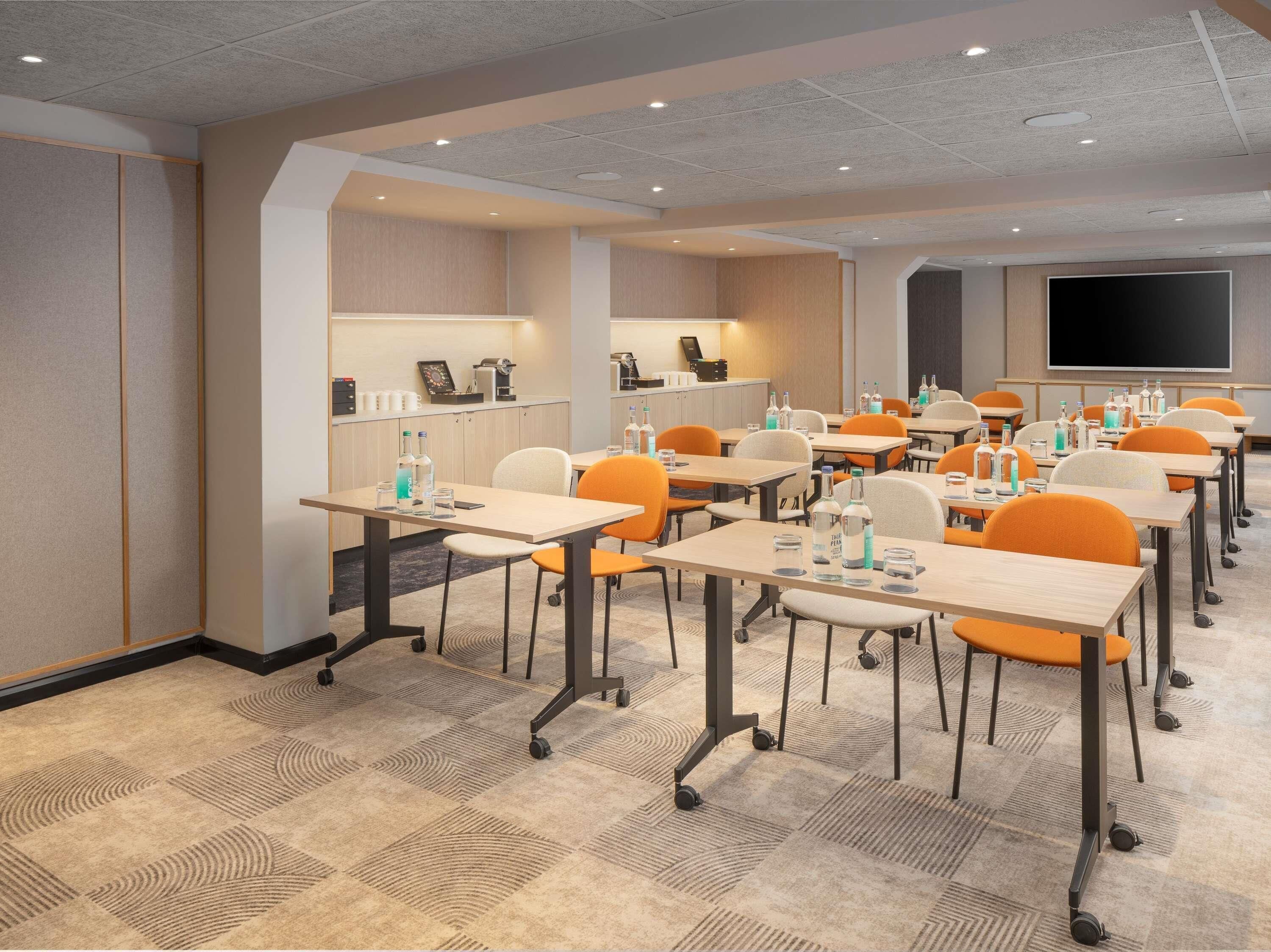 Sala de conferências com mesas, cadeiras, tela de projeção e um espaço de cozinha.