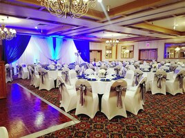 Salão de banquetes com mesas redondas, toalhas brancas e cadeiras decoradas com fitas.
