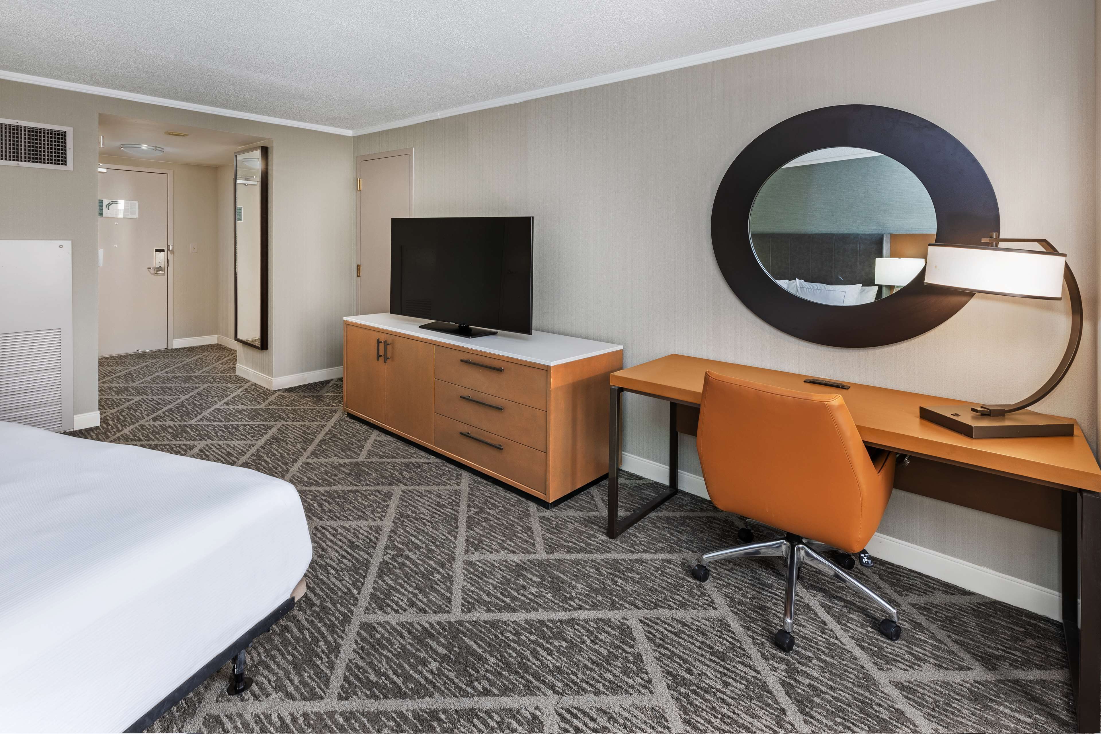 Quarto de hotel com cama, escrivaninha, cadeira, televisão, espelho e abajur.