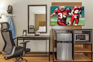 Quarto de hotel com escrivaninha, cadeira, frigorífico, micro-ondas e televisão exibindo uma partida de futebol.