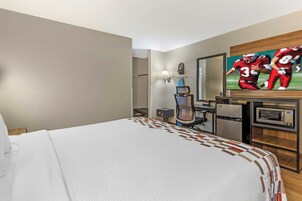 Quarto de hotel com uma cama grande, uma escrivaninha com computador, micro-ondas e televisão exibindo uma partida de futebol.