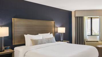 Quarto de hotel com uma cama grande, duas luminárias de cabeceira, um relógio digital e uma janela com cortinas.
