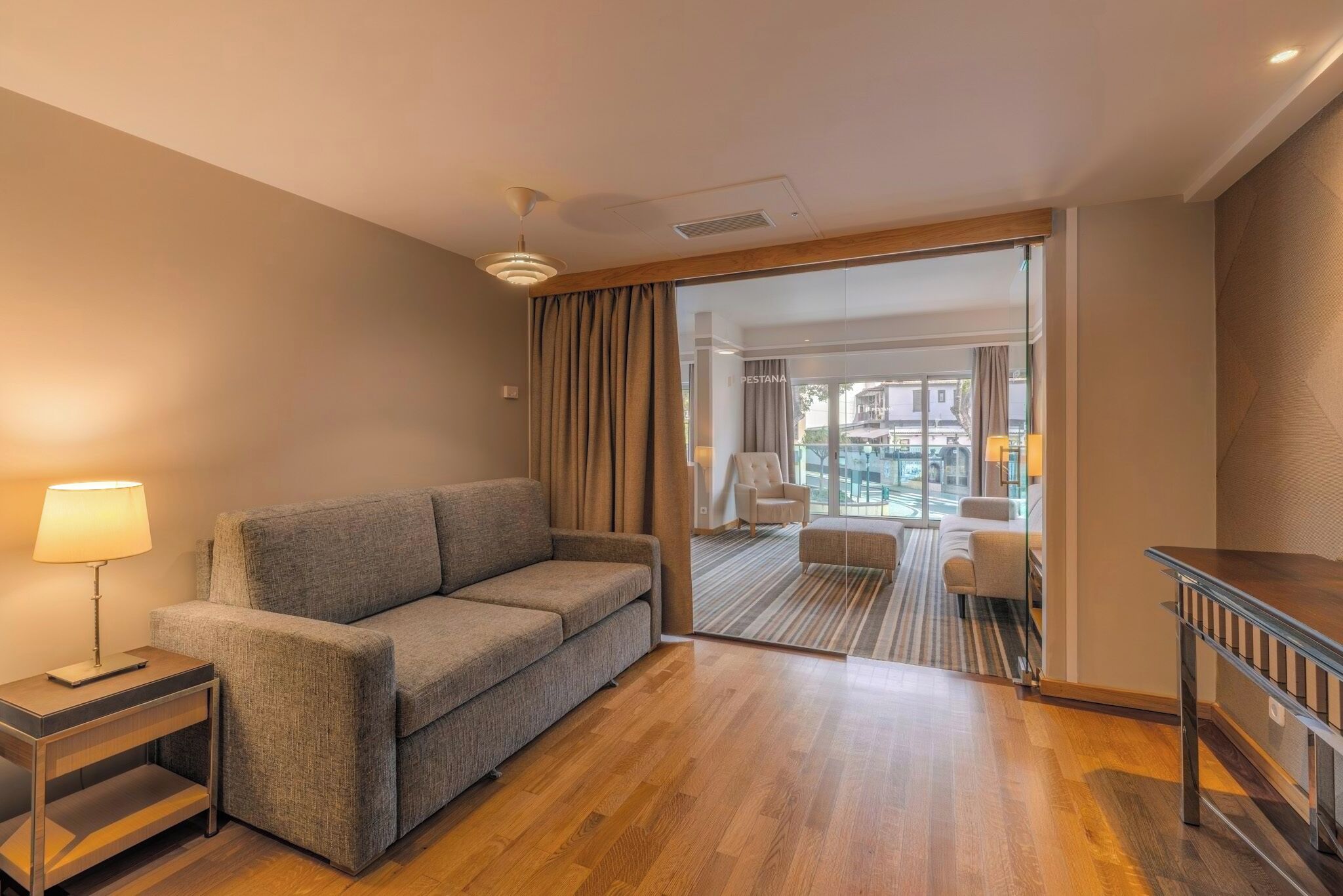 Quarto de hotel moderno com sofá, mesa lateral com abajur, piso de madeira e vista da cidade através de grandes janelas.