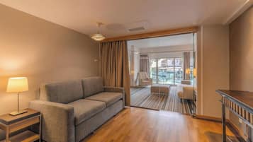 Quarto de hotel moderno com sofá, mesa lateral com abajur, piso de madeira e vista da cidade através de grandes janelas.