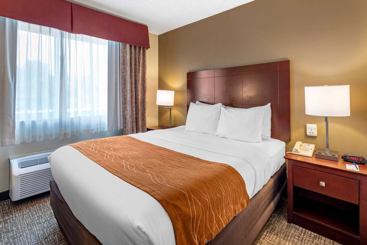 Quarto de hotel com uma cama grande, mesa de cabeceira com abajur, telefone e relógio digital.