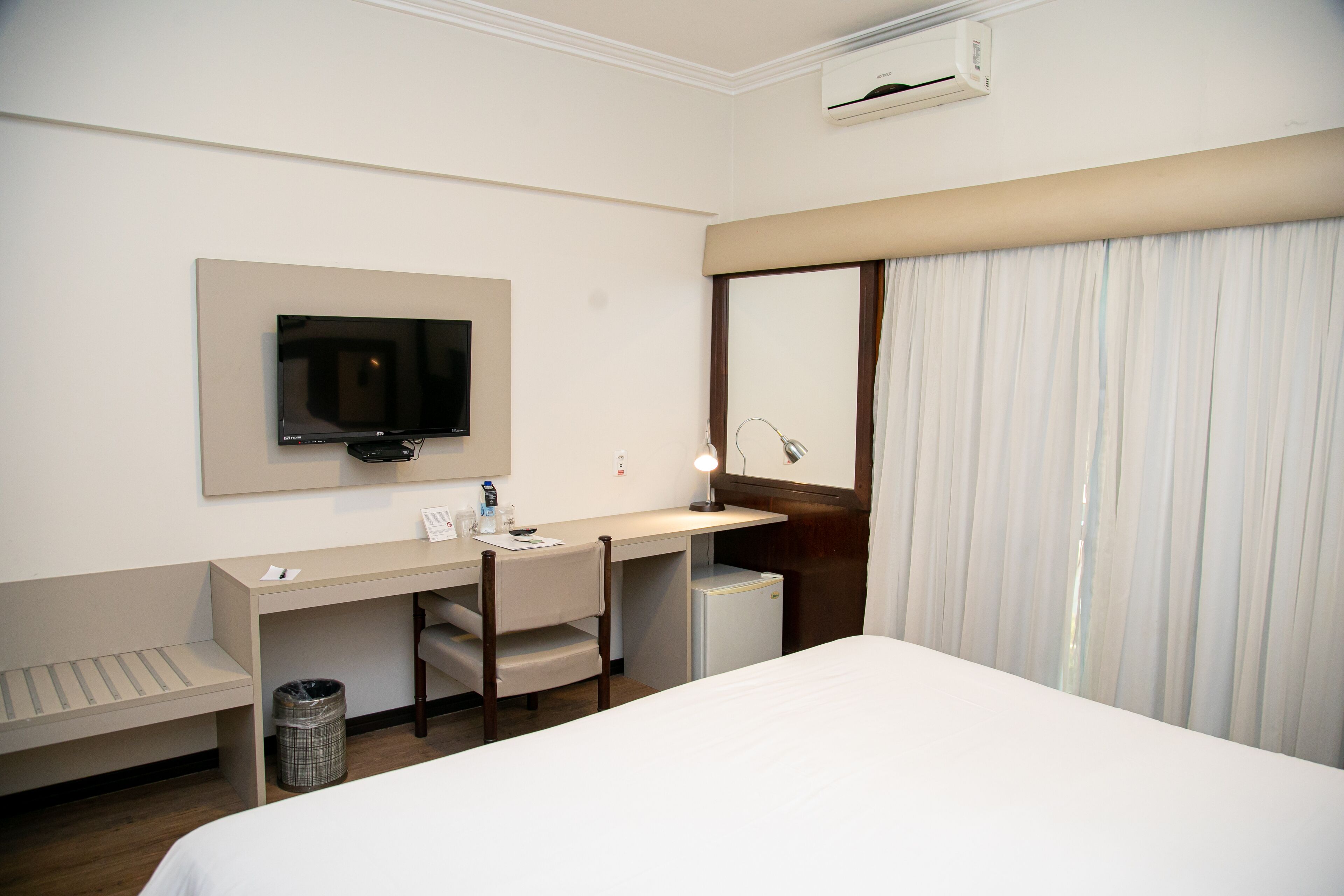 Quarto de hotel com cama, secretária com cadeira, televisão, espelho, pequeno frigorífico e lixeira.