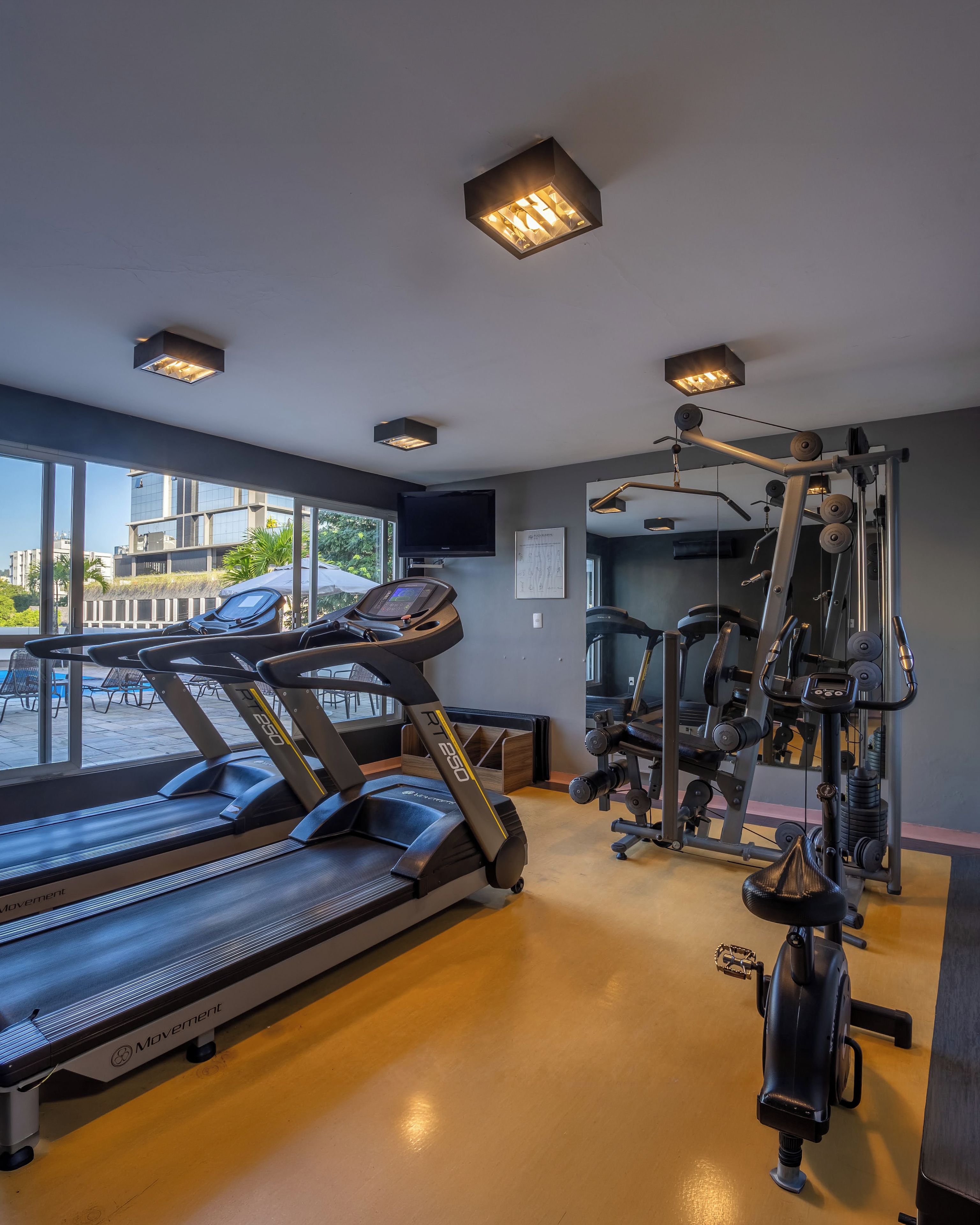 Academia com esteiras, máquinas de musculação e uma grande janela com vista para a paisagem urbana.