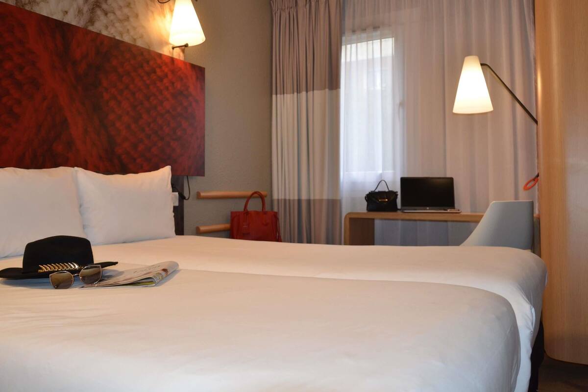 Quarto de hotel com uma cama grande, uma escrivaninha com um laptop, uma cadeira, uma bolsa vermelha e uma parede com textura.