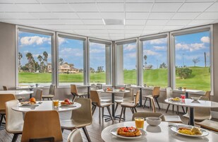 Uma área de refeições com grandes janelas que oferecem vista para um campo de golfe e um edifício. Mesas montadas com itens de café da manhã.