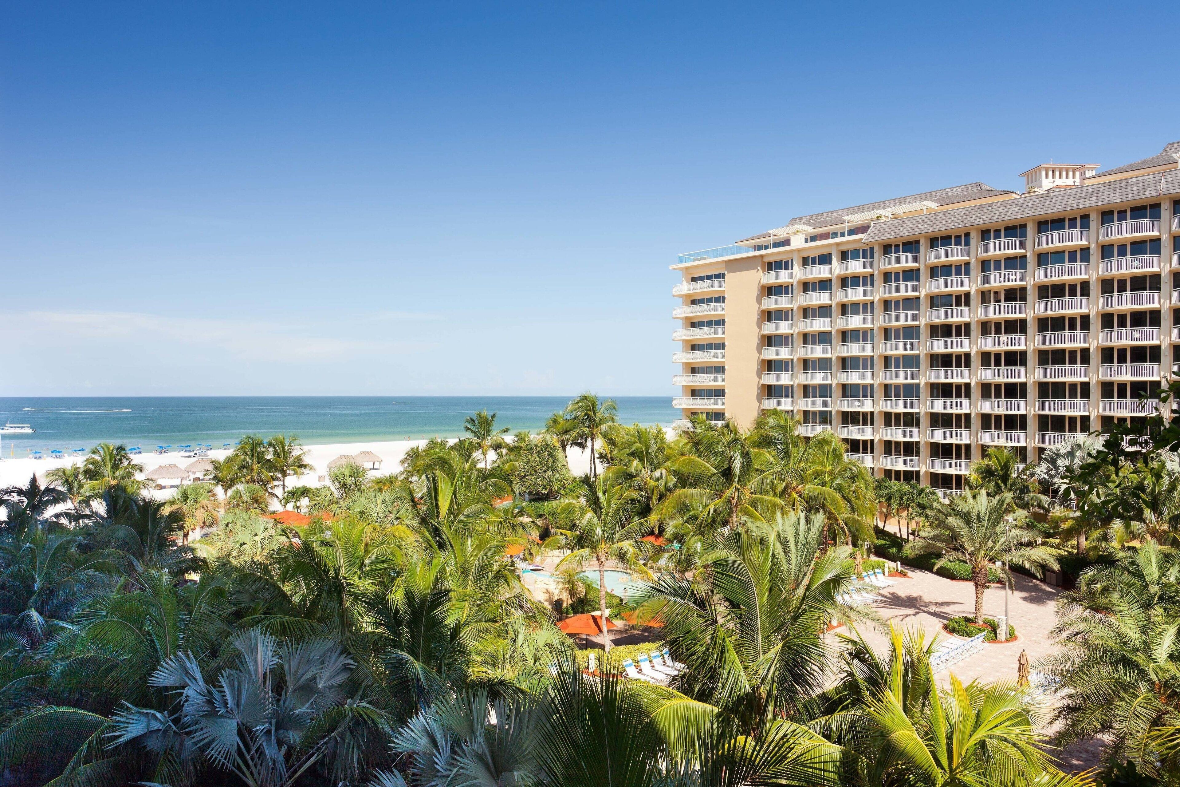 Um hotel com vista para a praia, várias varandas e uma área de piscina rodeada de palmeiras.