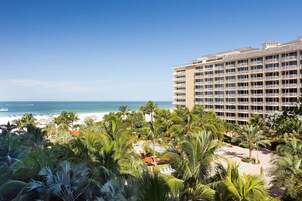 Um hotel com vista para a praia, várias varandas e uma área de piscina rodeada de palmeiras.