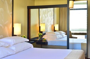 Quarto de hotel com uma cama grande, uma mesa de cabeceira com luminária, um espelho e vista para o mar.