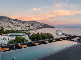 Piscina infinita no terraço com espreguiçadeiras, vista de um navio de cruzeiro e paisagem urbana com colinas ao fundo.