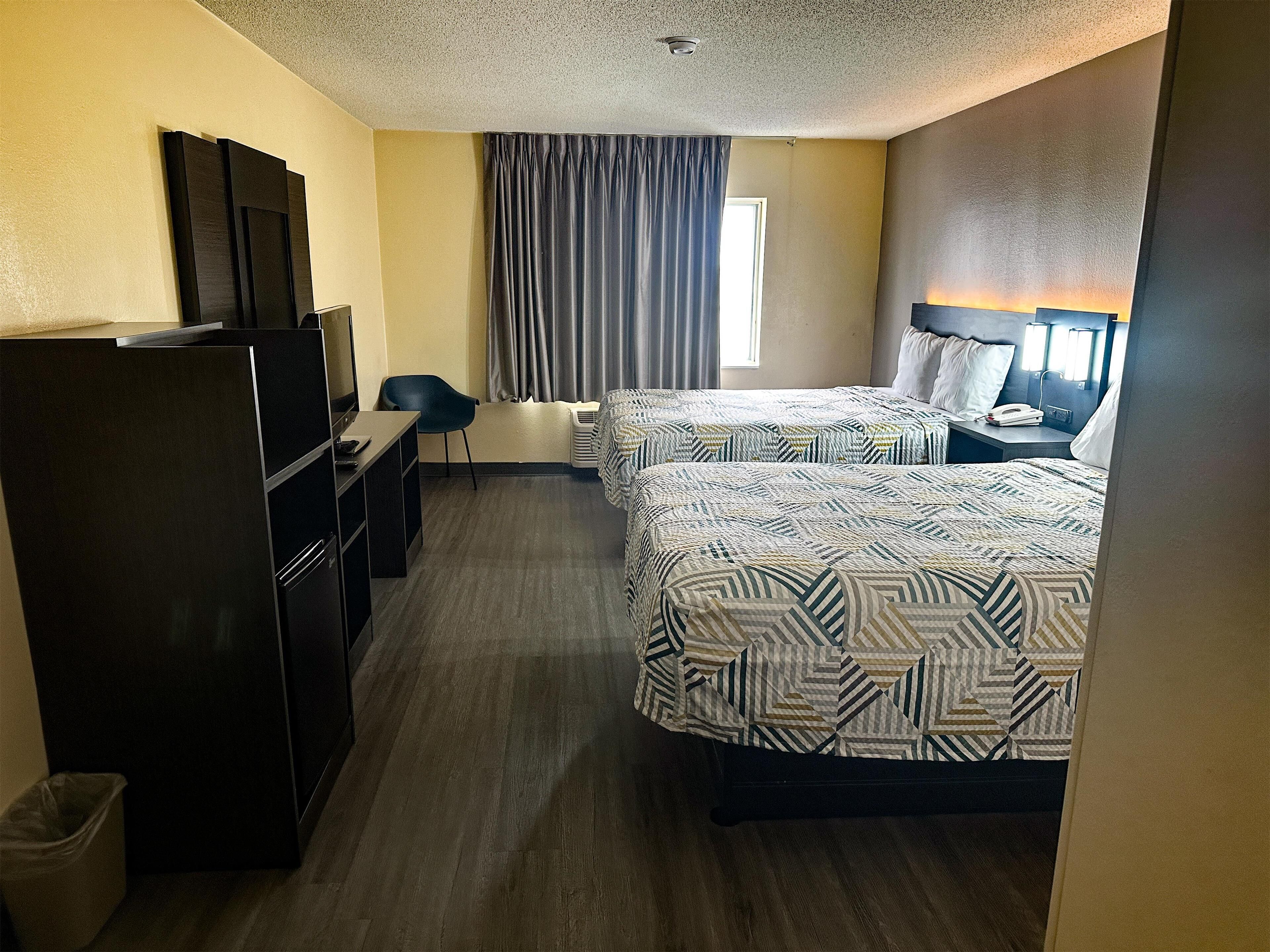 Quarto de hotel com duas camas, uma mesa de trabalho com televisão, uma cadeira, uma janela com cortinas e uma luminária.
