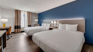 Quarto de hotel com duas camas, uma escrivaninha, uma cadeira e uma luminária.