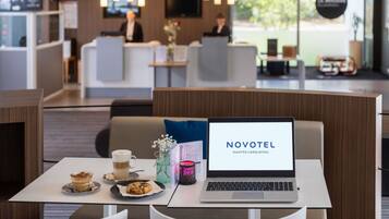 Área de recepção de um hotel Novotel, com um laptop sobre uma mesa, uma chávena de café, um biscoito e um vaso com flores.