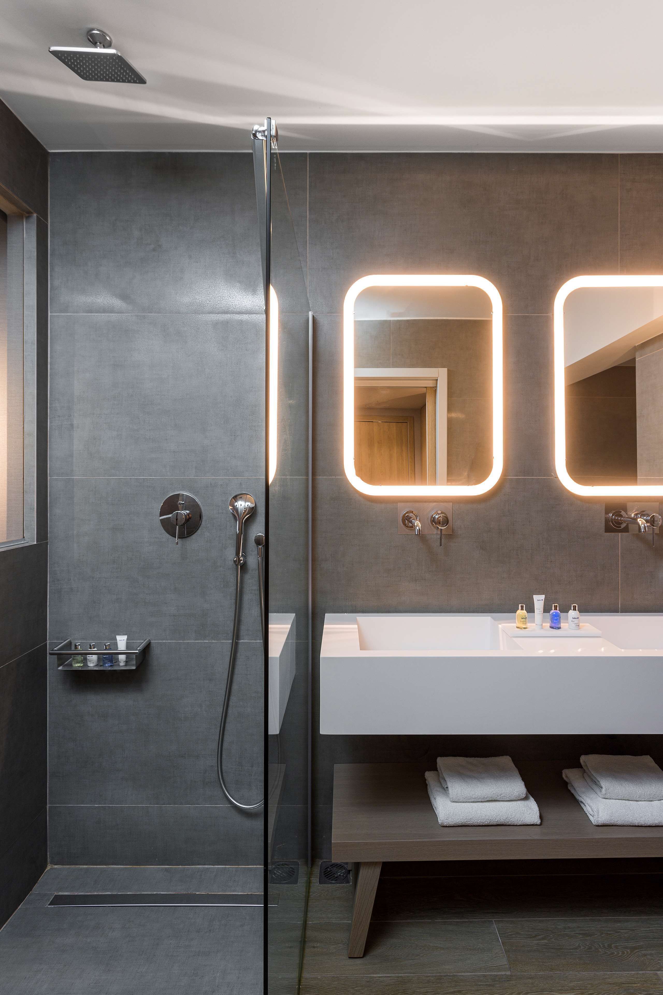 Uma casa de banho moderna com cabine de duche em vidro, dois espelhos com iluminação e toucador com lavatório e espaço para toalhas.