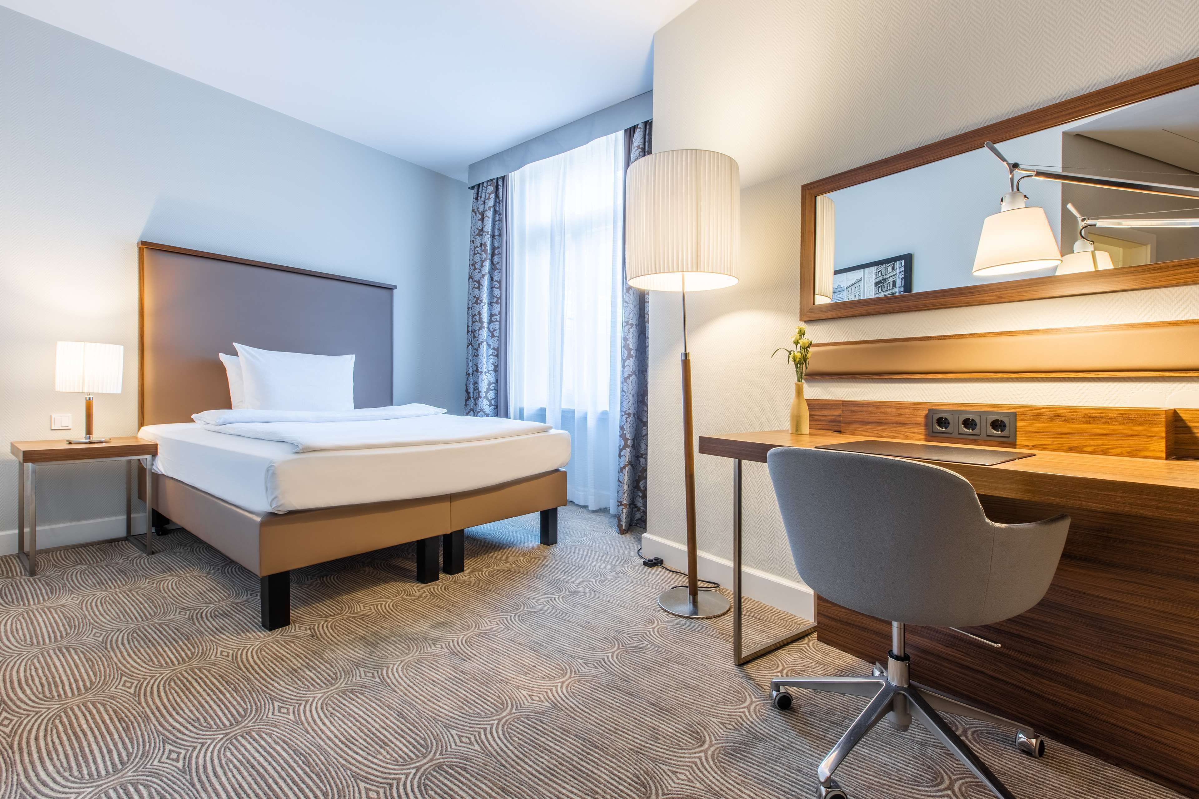Quarto de hotel com cama, escrivaninha, cadeira, luminária e espelho.