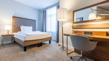 Quarto de hotel com cama, escrivaninha, cadeira, luminária e espelho.