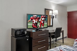 Quarto de hotel com televisão exibindo uma partida esportiva, escrivaninha com cadeira, micro-ondas e armário com gavetas.
