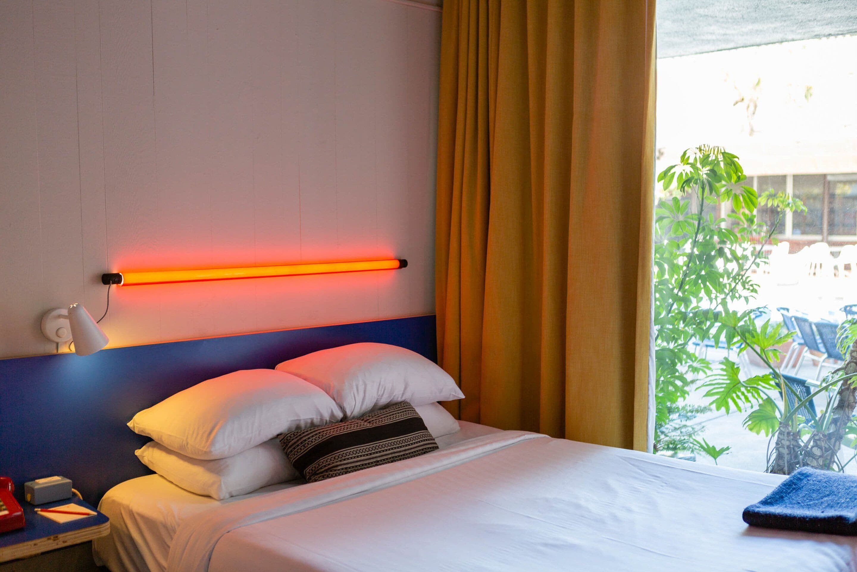 Quarto de hotel com uma cama, mesa de cabeceira, luminária e janela com cortinas.