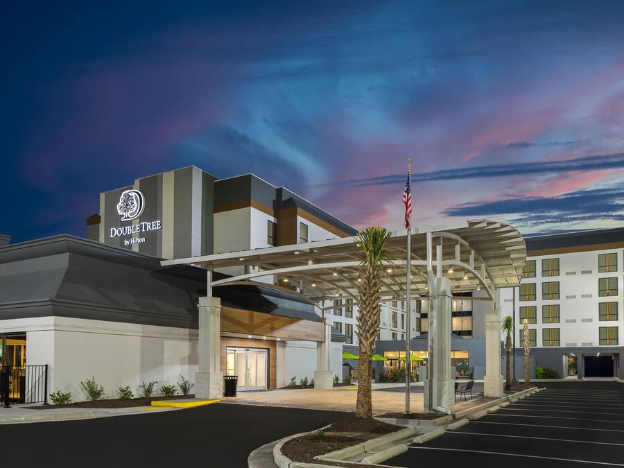 Hotel com design moderno, destacando uma entrada imponente com cobertura e um mastro.