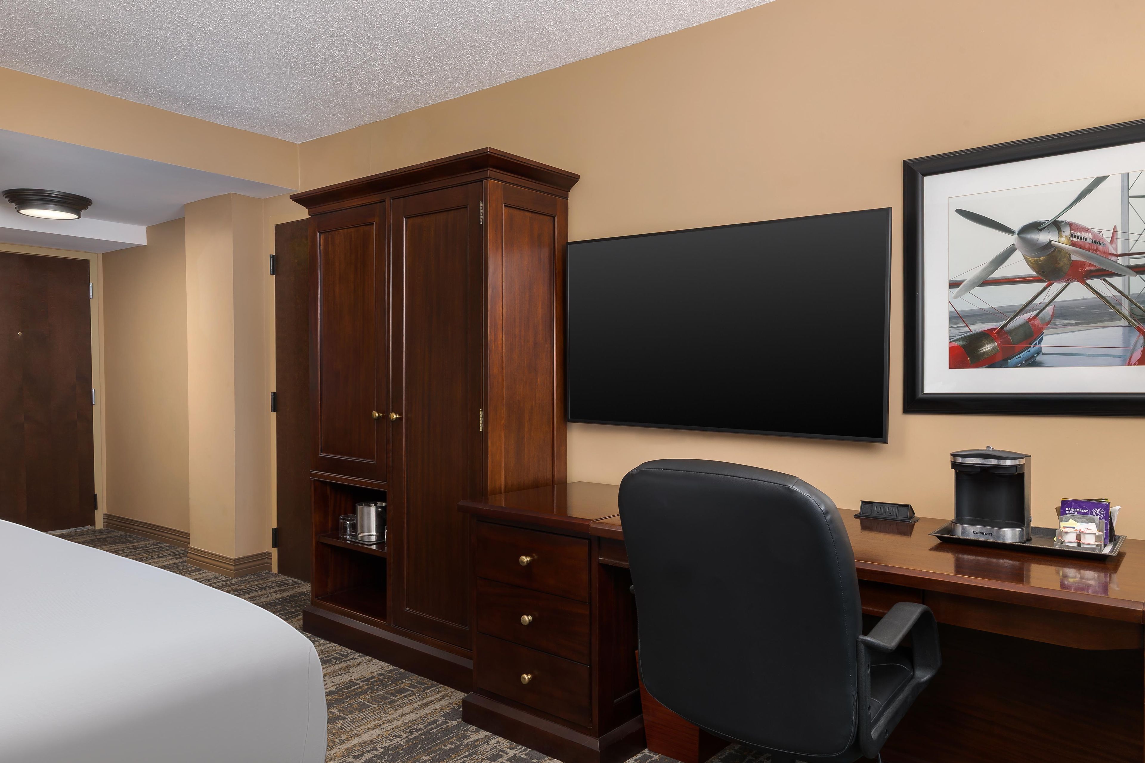 Quarto de hotel com guarda-roupa de madeira grande, escrivaninha com cadeira, televisão de tela plana e quadro com fotografia de um avião.