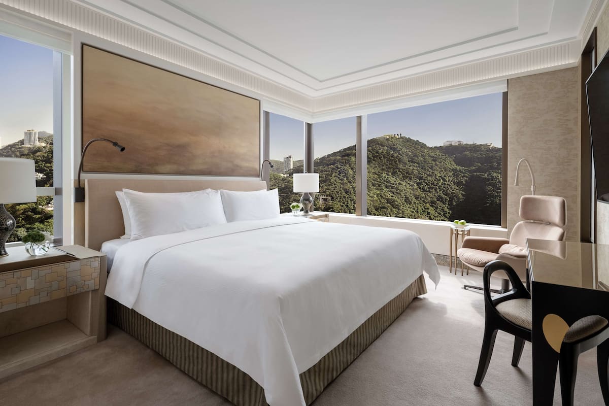 Quarto de hotel com uma cama grande, uma mesa com cadeira, vista para uma paisagem montanhosa e um quadro grande na parede.
