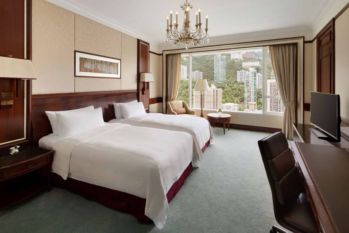 Quarto de hotel com duas camas, uma televisão, escrivaninha, cadeira, lustre e uma grande janela com vista para a cidade.