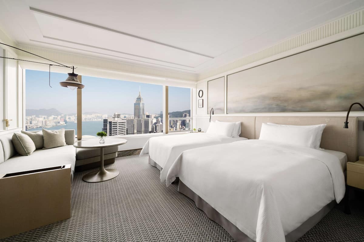 Quarto de hotel com duas camas, um sofá, uma mesa redonda e uma grande janela com vista para a cidade.