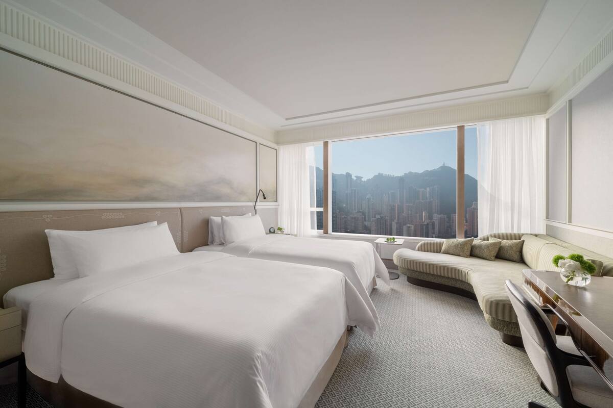 Quarto de hotel com duas camas, um sofá, uma escrivaninha e uma janela grande com vista para a cidade.