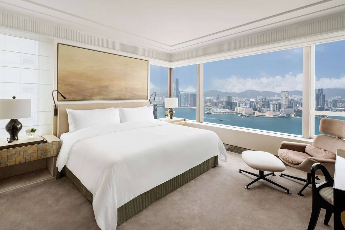 Quarto de hotel com uma cama grande, uma escrivaninha com cadeira e vista para a paisagem urbana através de amplas janelas.