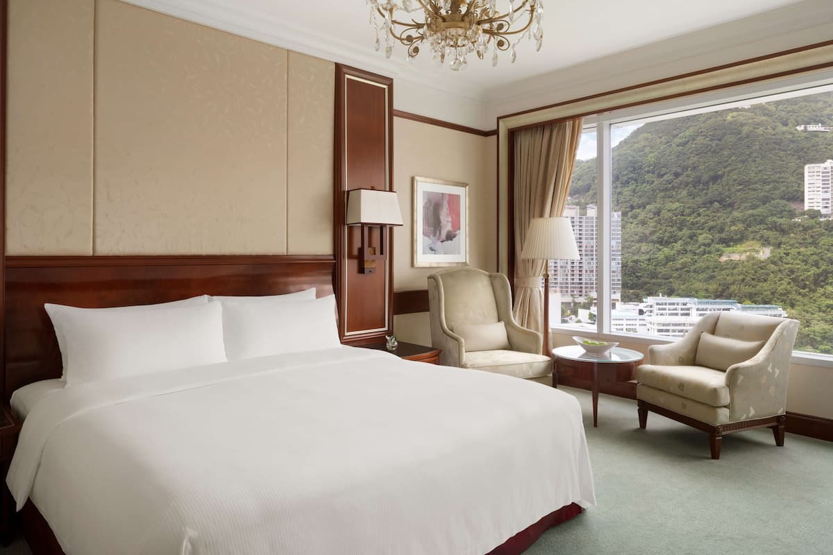 Quarto de hotel com uma cama grande, uma cadeira, uma mesinha e vista para o horizonte urbano.
